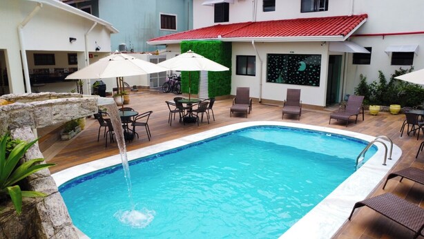 Piscina com muro de pedra, rodeada por espreguiçadeiras e guarda-sóis, e um edifício com telhado vermelho ao fundo.
