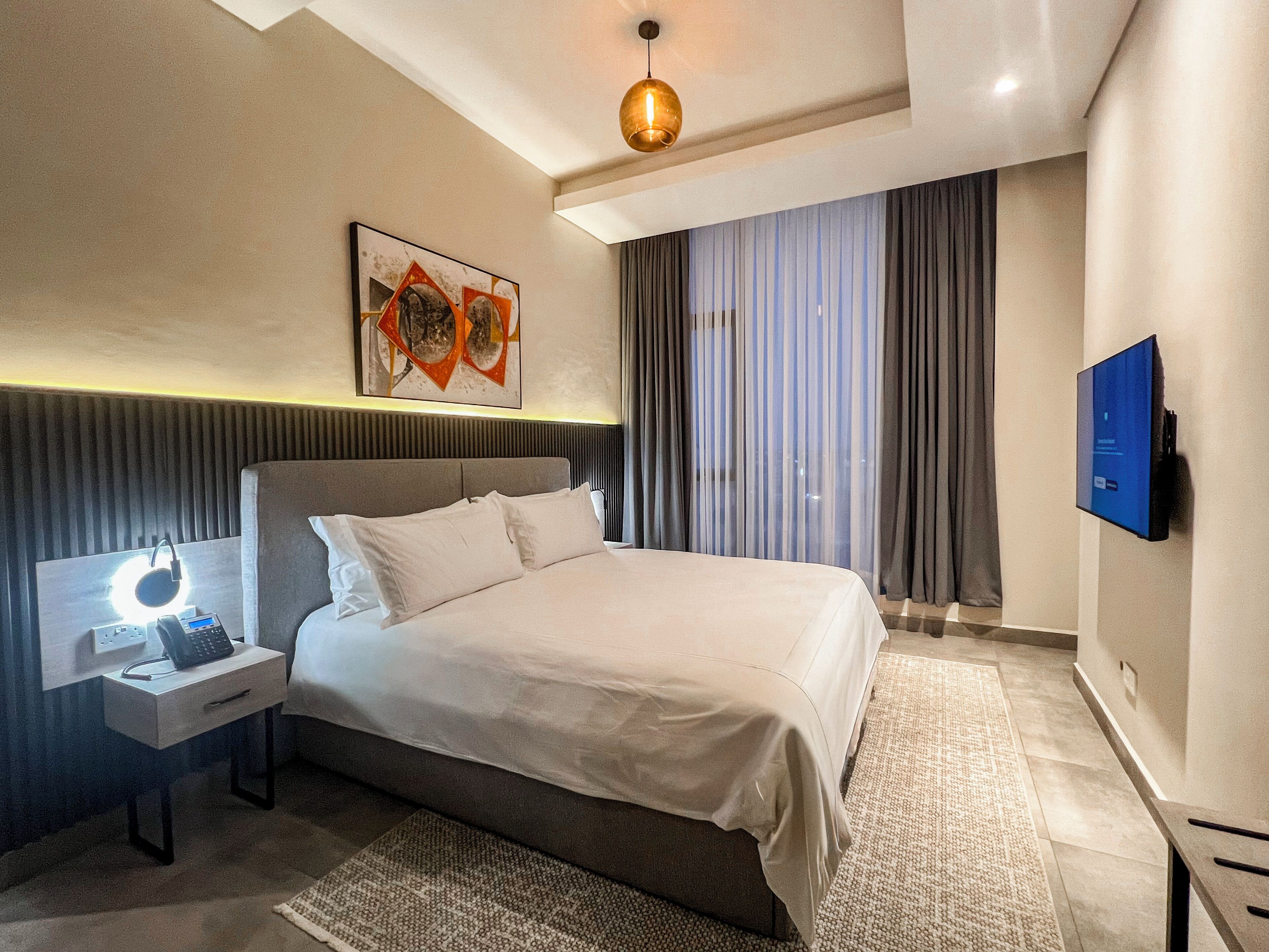 Quarto de hotel moderno com uma cama grande, mesa de cabeceira com abajur, televisão de tela plana na parede e janela com cortinas.