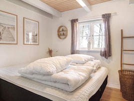 Cama bem arrumada com colchão acolchoado e almofadas, janela com cortinas e prateleira fixa na parede com objetos decorativos.