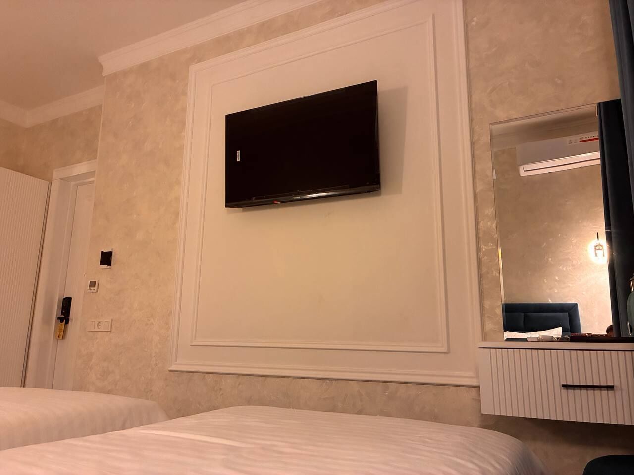 Quarto de hotel com televisão de tela plana na parede, mesa de cabeceira com espelho e duas camas.