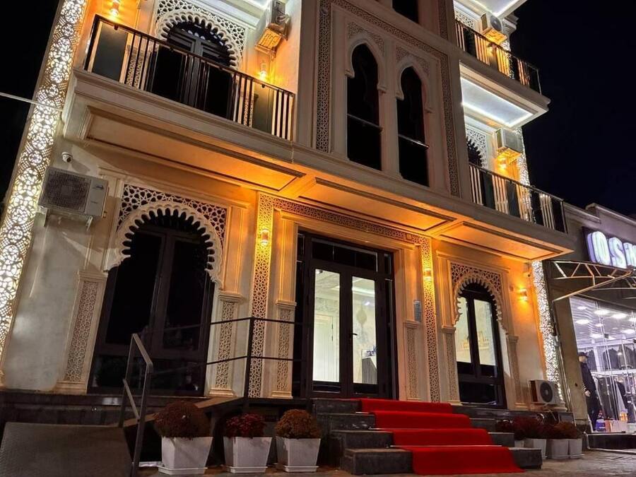 Um edifício de dois andares com detalhes arquitetônicos ornamentados, incluindo arcos e colunas, e um tapete vermelho que conduz à entrada.