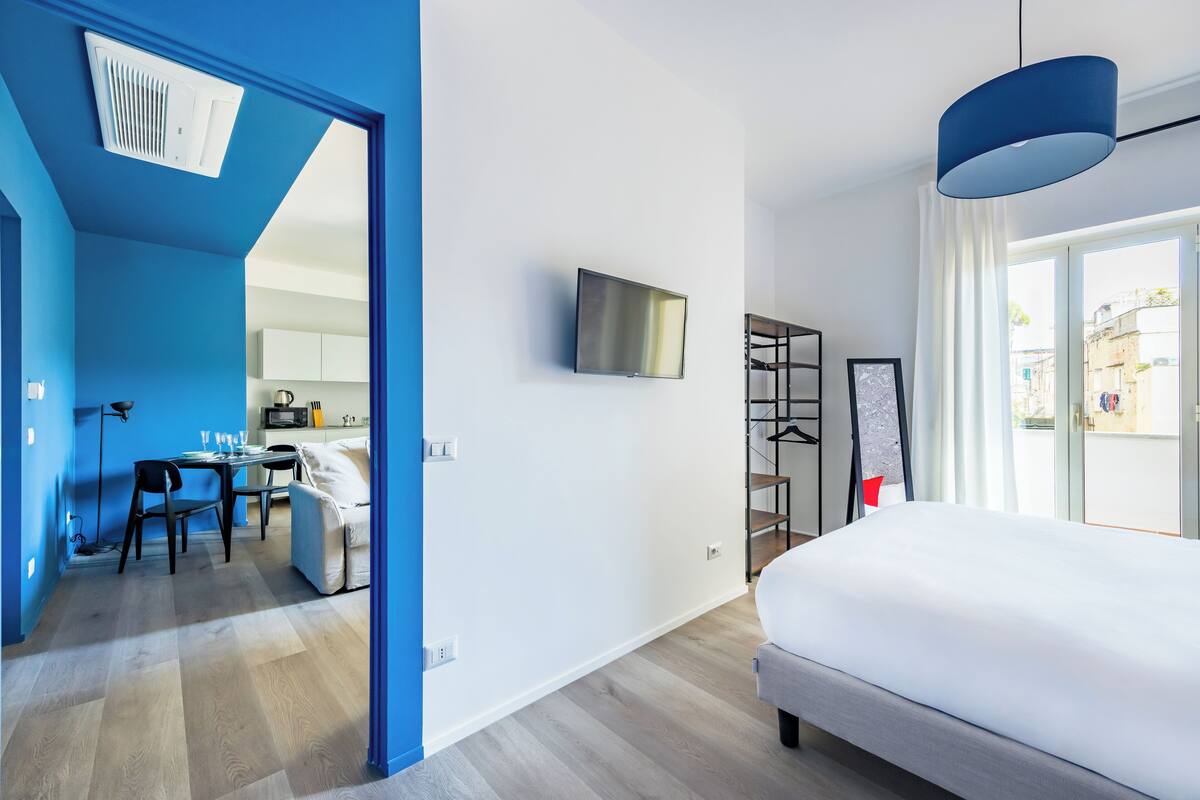 Quarto de hotel moderno com uma cama grande, televisão de tela plana e uma área de refeições visível através de uma porta aberta.