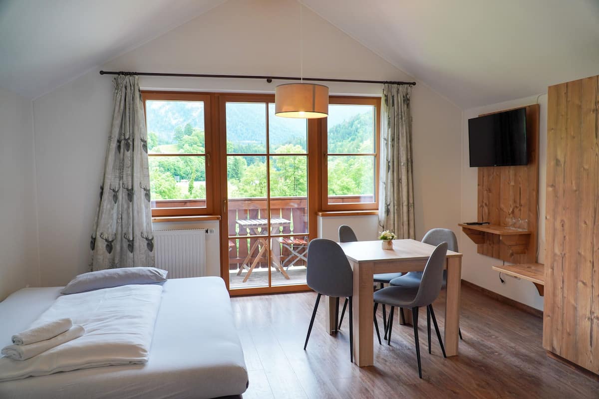 Quarto com cama, mesa e cadeiras, TV fixada em um painel de madeira, e vista para o verde através de grandes janelas.