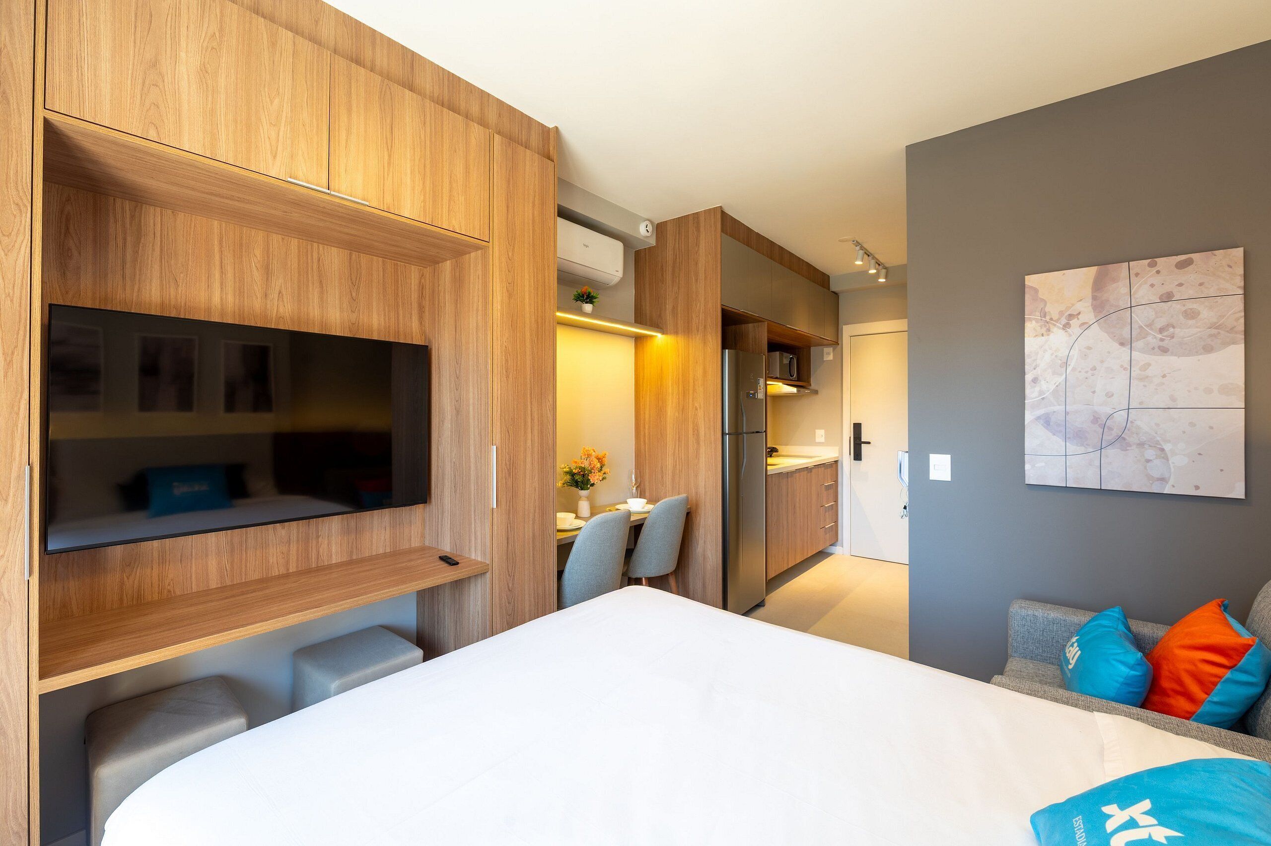 Quarto de hotel moderno com uma cama grande, armários de madeira, área de refeições e cozinha.