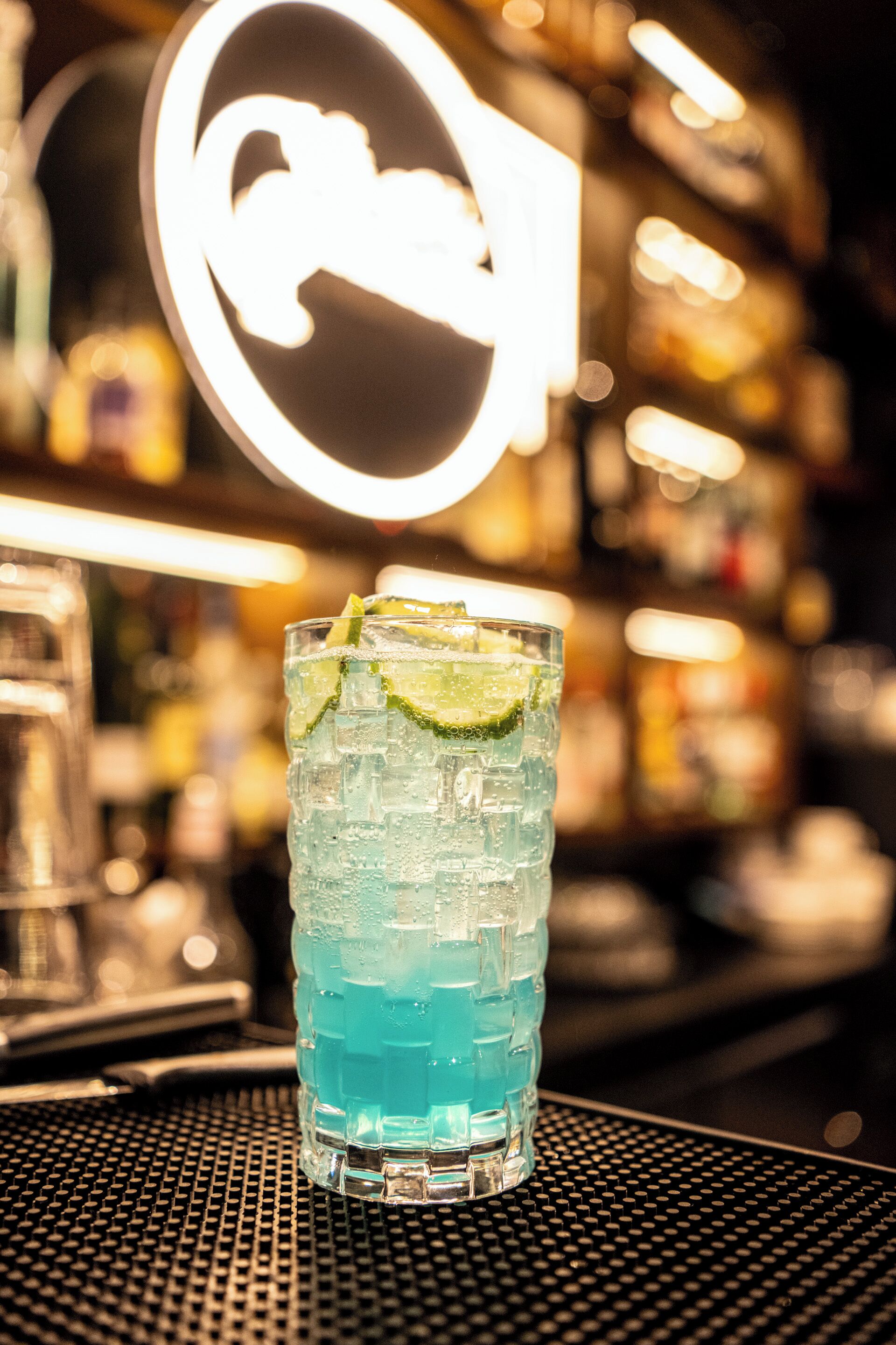 Um bar, com um copo de bebida azul decorado com limão, uma placa de neon ao fundo e um balcão.