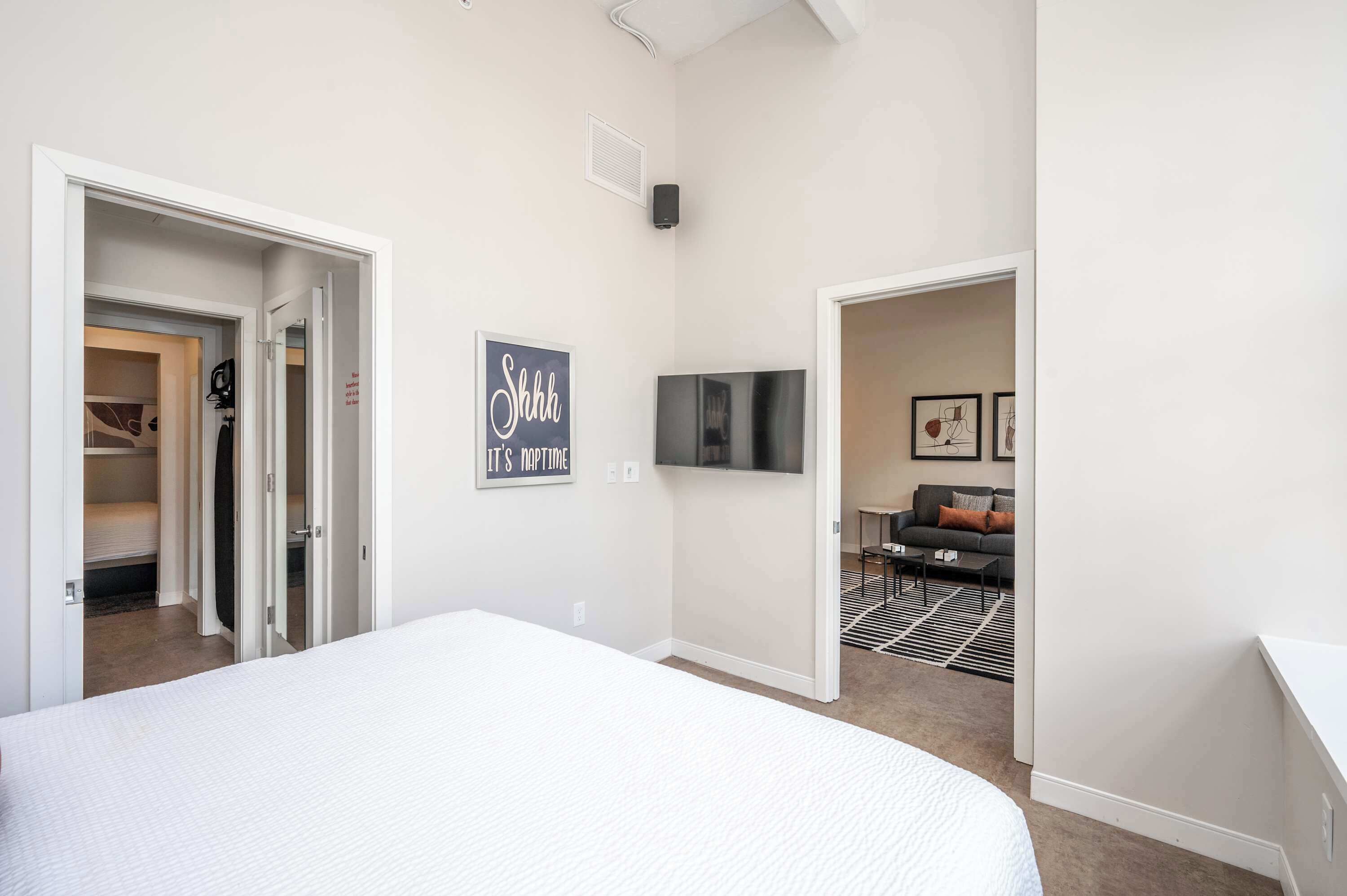 Quarto com uma cama grande, televisão presa na parede e corredor que leva a uma sala de estar com sofá e obras de arte.