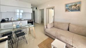 Uma sala de estar moderna com um sofá bege, uma mesa de jantar de vidro com cadeiras, uma área de cozinha com armários brancos e um quadro na parede.