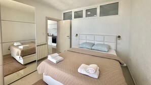 Quarto moderno com uma cama grande, um guarda-roupa espelhado e uma pequena cozinha compacta ao fundo.