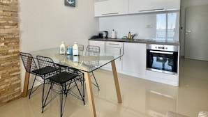 Uma cozinha moderna com armários brancos, forno de aço inoxidável e uma área de jantar com mesa de vidro e cadeiras de metal.