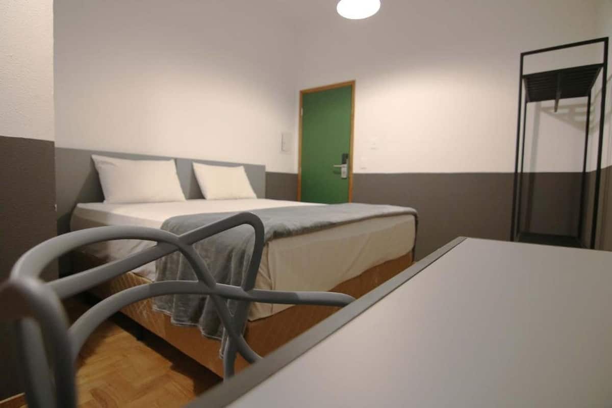 Uma cama de solteiro com cabeceira cinza, uma porta verde e uma divisória de vidro à direita.