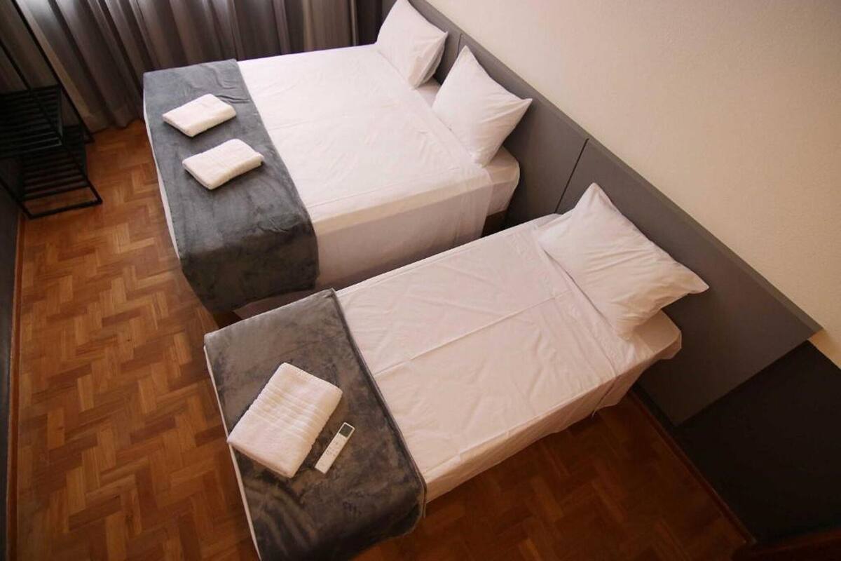 Quarto de hotel com duas camas de solteiro, uma mesinha com um livro e um controle remoto, e piso de madeira.