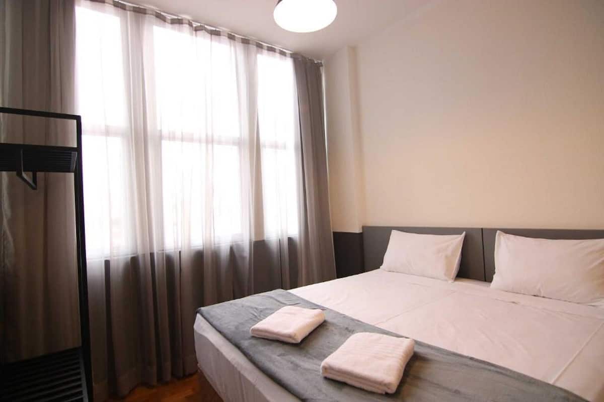 Quarto de hotel com cama, duas almofadas, mesa de cabeceira com toalhas e janela com cortinas.