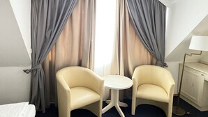 Quarto de hotel com cama de canto, duas poltronas, uma mesinha redonda e uma luminária.