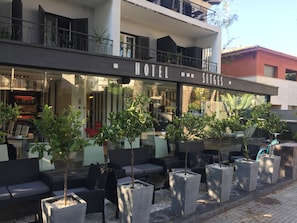 Hotel com área externa para sentar, plantas em vasos e vista clara do interior através da fachada de vidro.