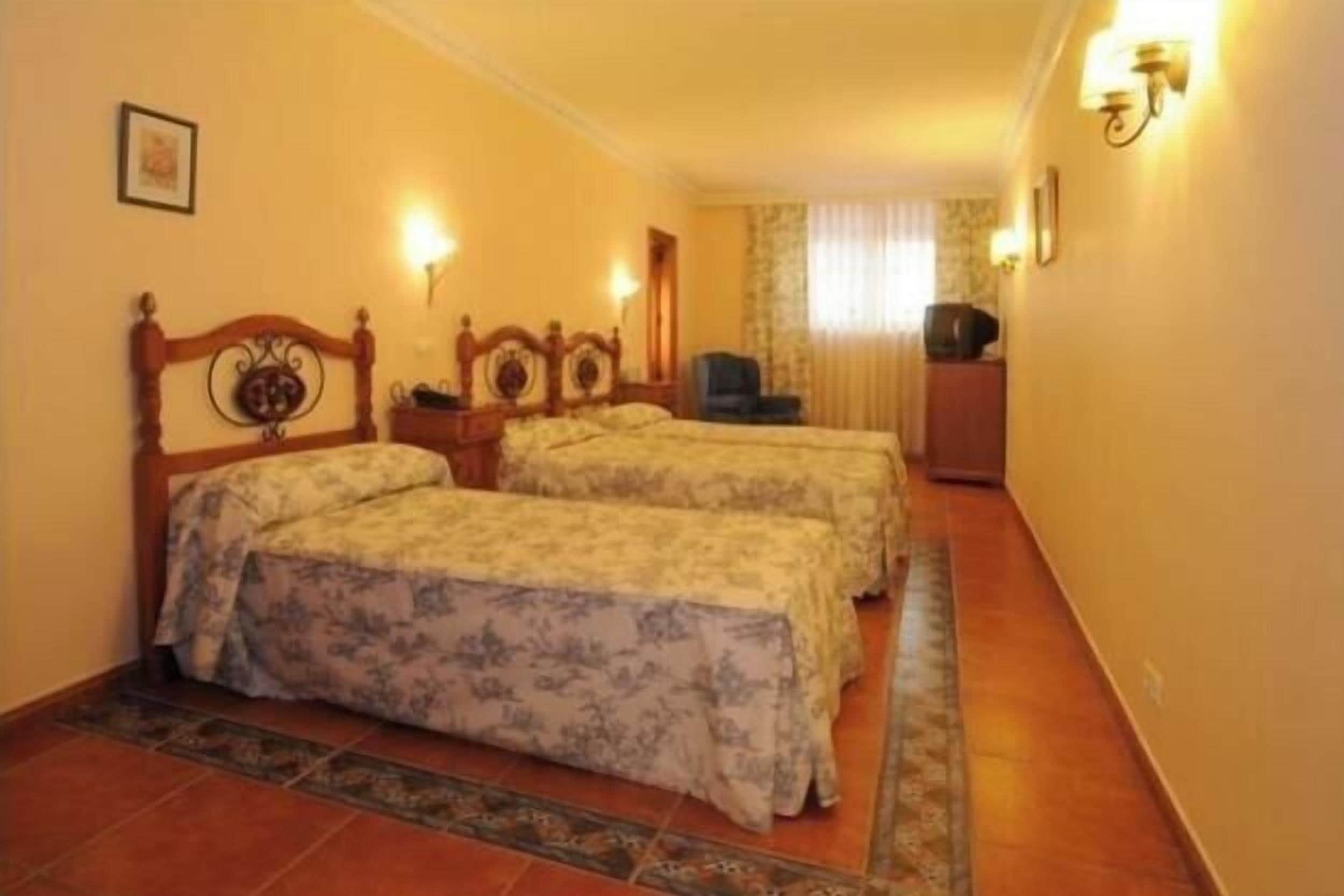 Quarto de hotel com duas camas, uma pequena escrivaninha, uma cadeira e uma janela com cortinas.