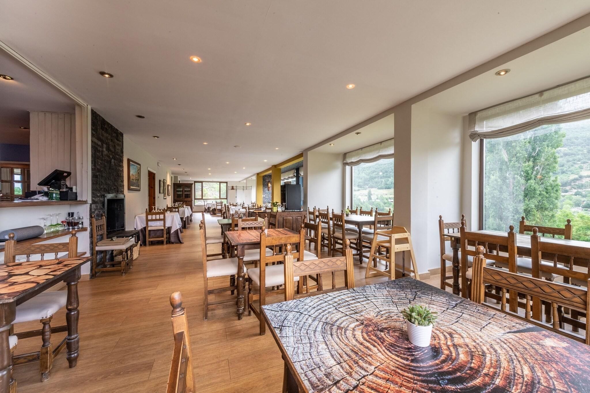 Uma ampla sala de jantar com várias mesas e cadeiras de madeira, uma parede decorada com pedras e grandes janelas com vista para o exterior.