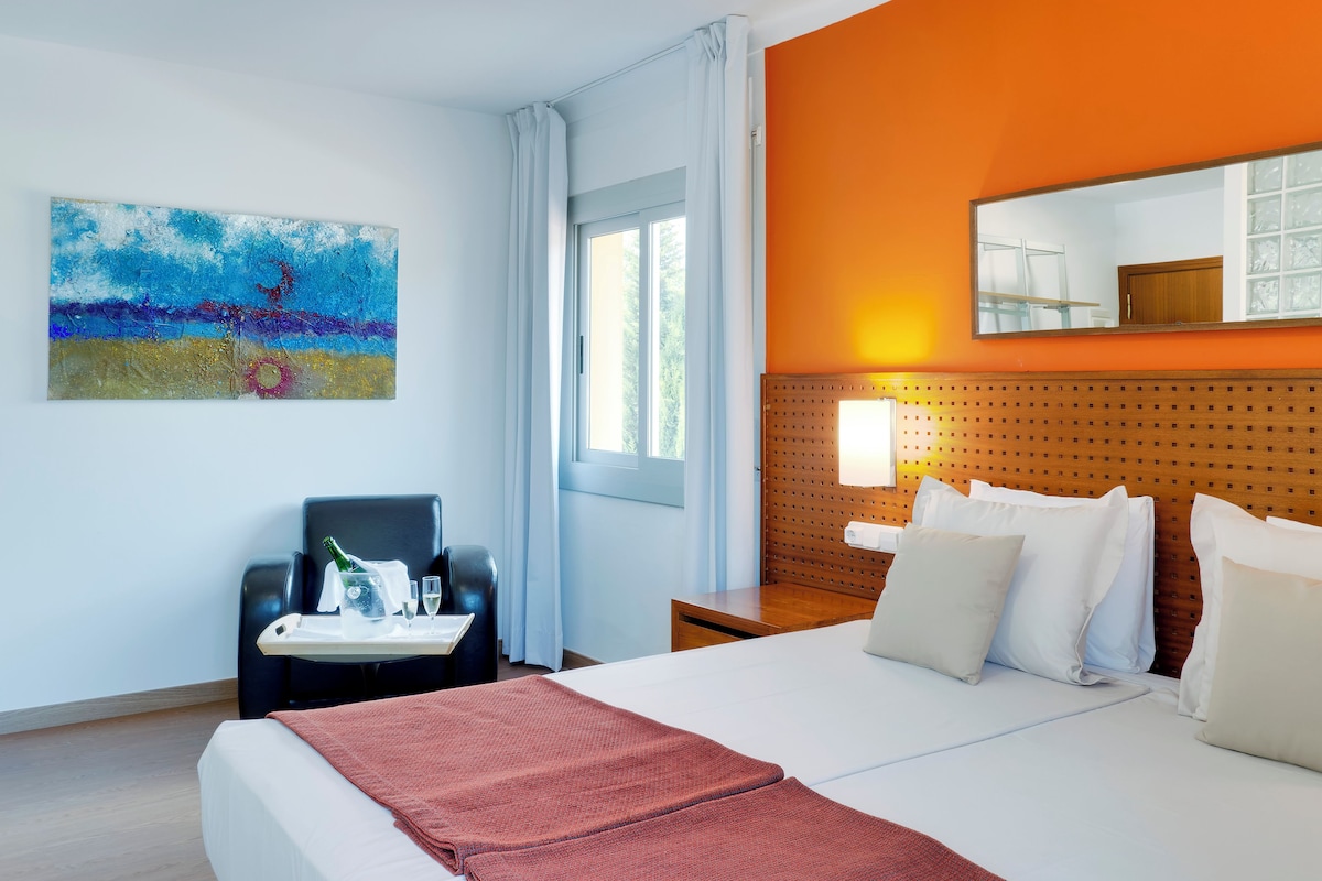 Quarto de hotel com cama, escrivaninha com cadeira, um quadro e uma janela.