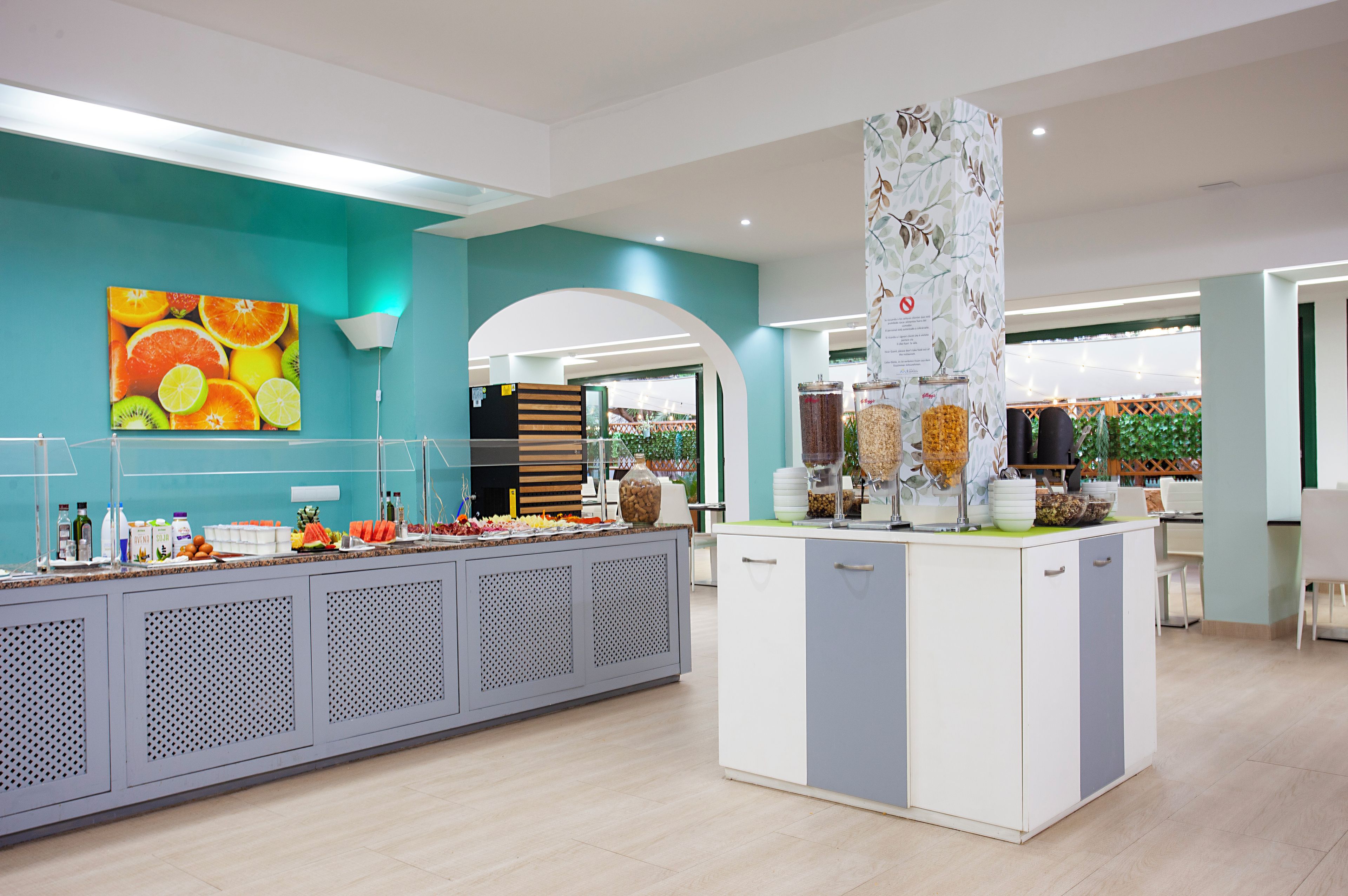 Uma área de café da manhã moderna, com uma variedade de frutas, um dispensador de sucos e uma estação de café.