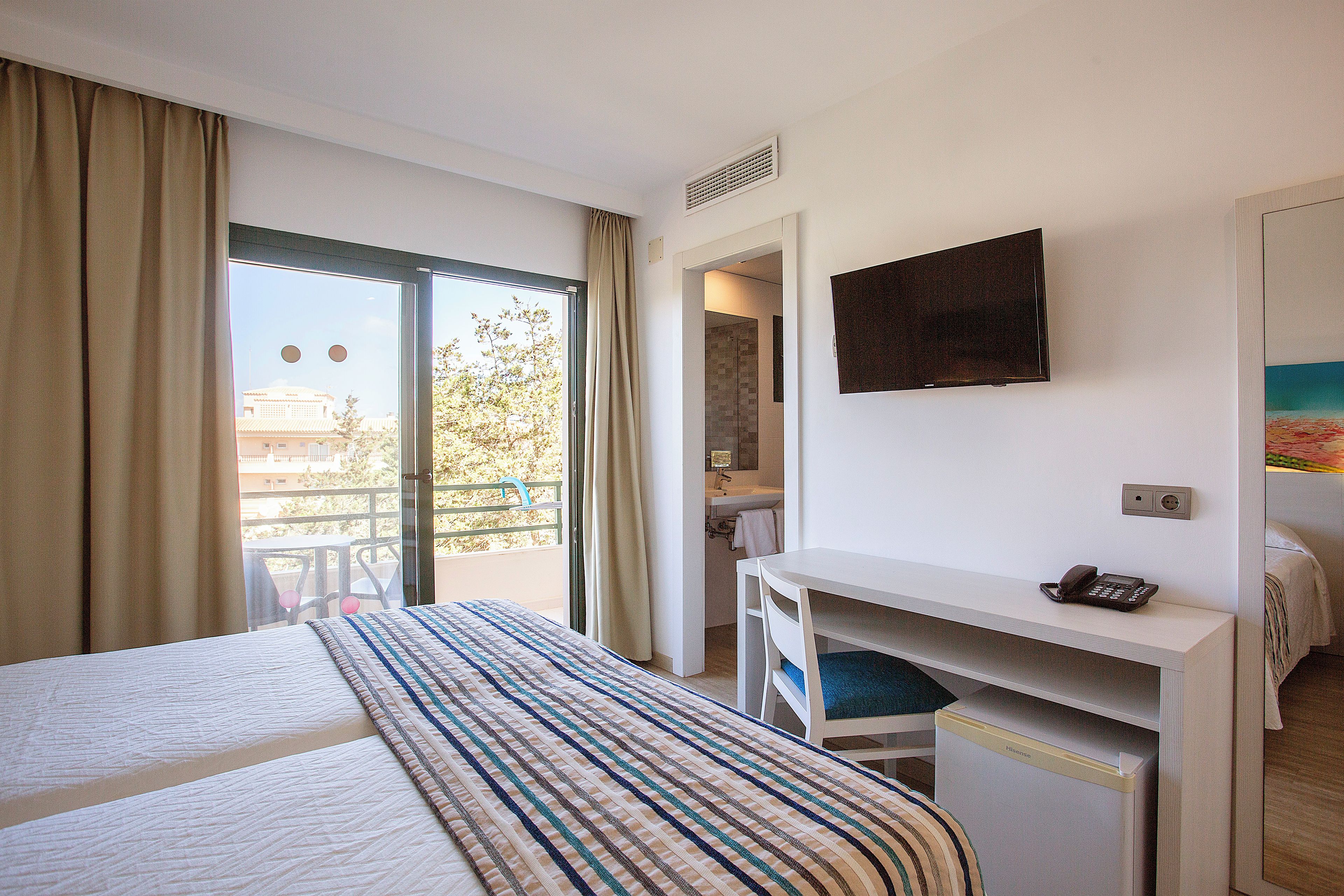 Quarto de hotel com uma cama, uma secretária com cadeira, televisão e banheiro visível por uma porta aberta.