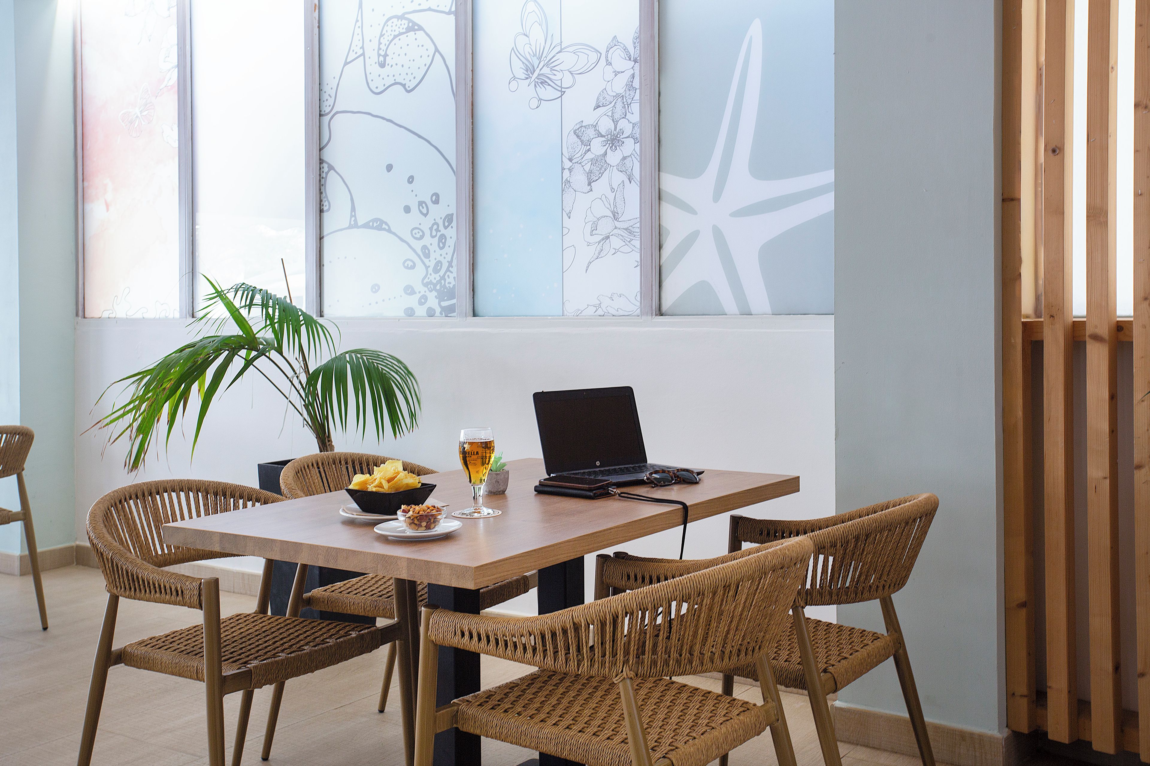 Uma sala de jantar com uma mesa de madeira, cadeiras de vime, um laptop, um copo de cerveja e uma planta.