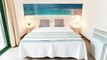 Quarto de hotel com uma cama, duas luminárias de cabeceira, uma mesinha de apoio, uma janela com cortinas e uma TV fixada na parede.