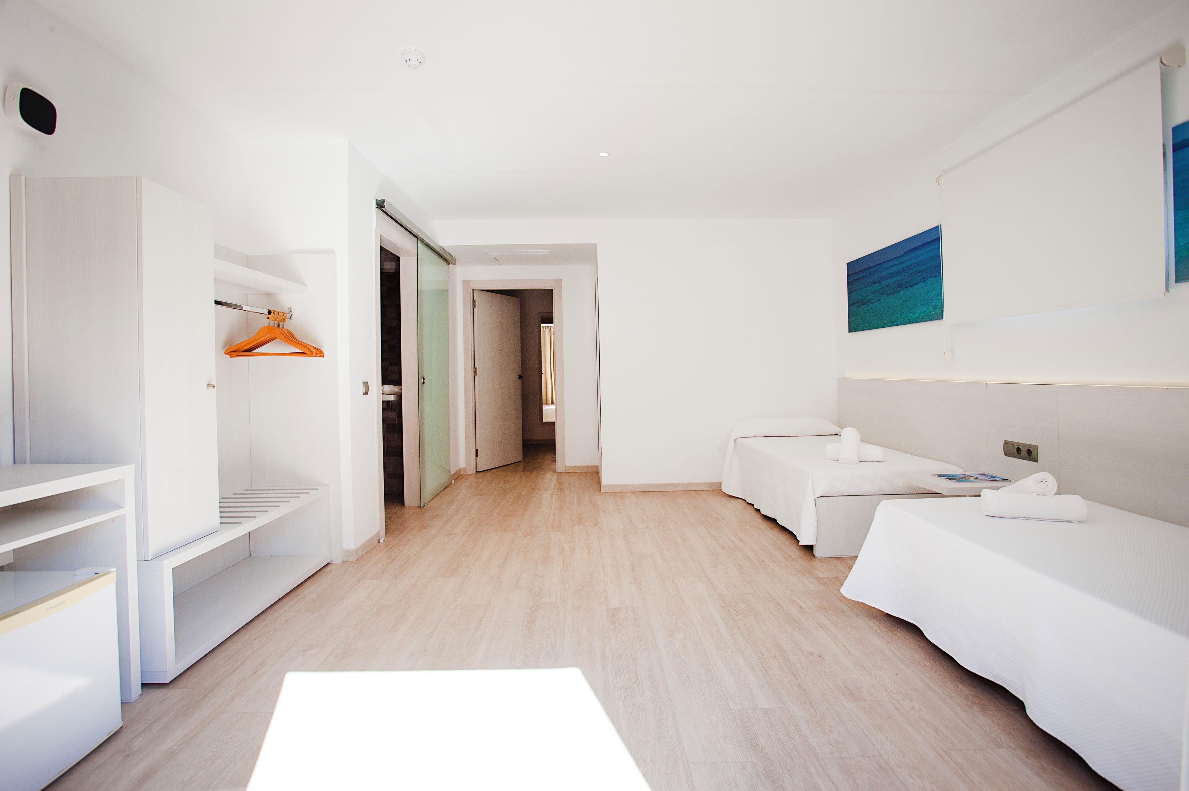 Quarto de hotel moderno com piso de madeira, móveis brancos, uma cama, um espaço de estar e um corredor que leva a outro quarto.