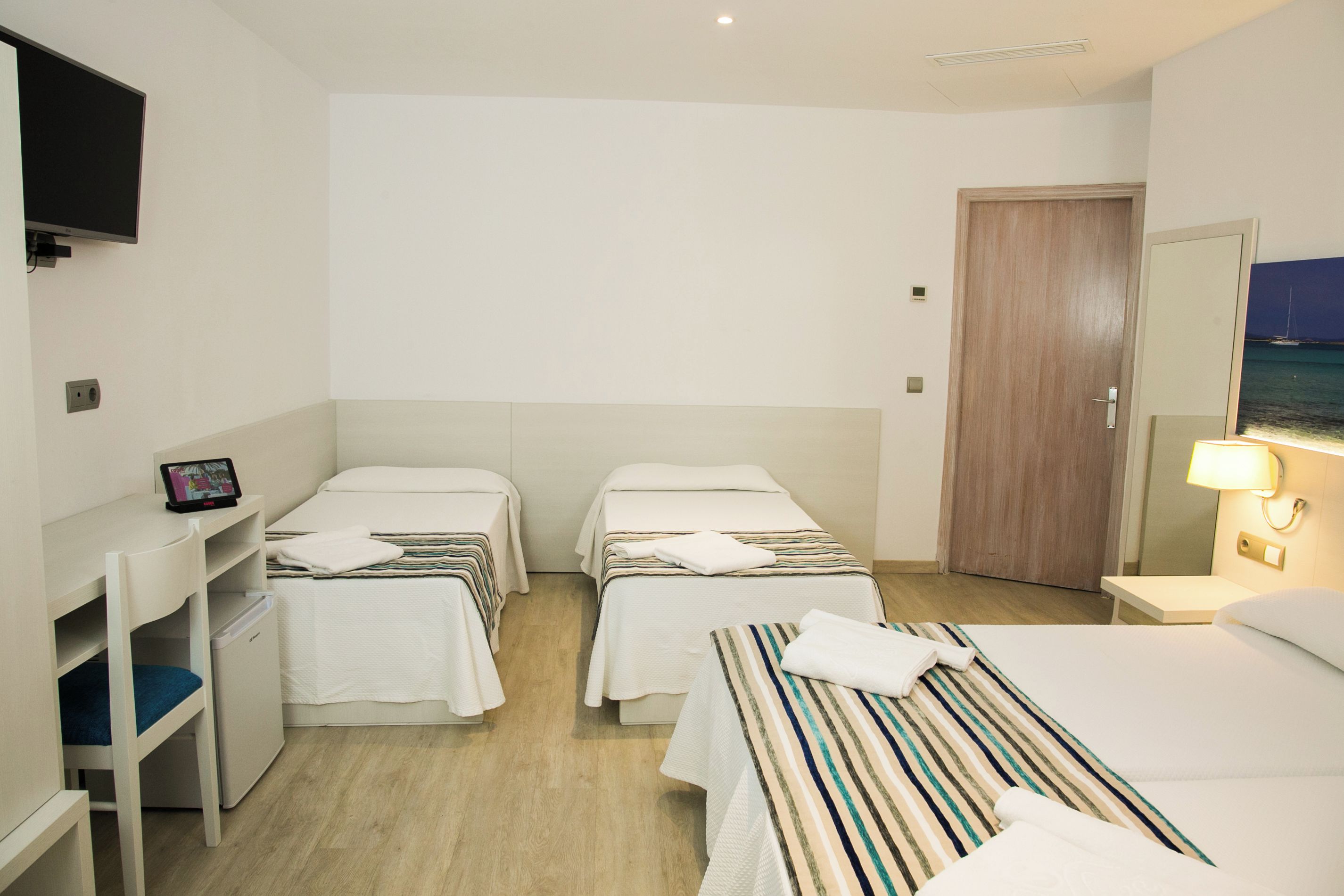 Quarto de hotel com duas camas de solteiro, uma mesa com cadeira, TV fixada na parede, porta e mesa de cabeceira com abajur.