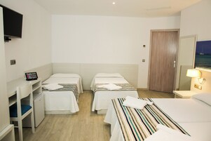 Quarto de hotel com duas camas de solteiro, uma mesa com cadeira, TV fixada na parede, porta e mesa de cabeceira com abajur.