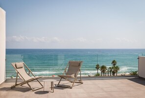 Varanda com duas cadeiras de descanso e uma mesa pequena, com vista para uma praia com palmeiras e o oceano.
