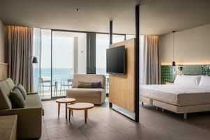 Quarto de hotel com cama, sofá, televisão, mesinha e varanda vista para o mar.