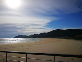 Uma praia com céu claro, montanhas ao fundo e uma grade em primeiro plano.