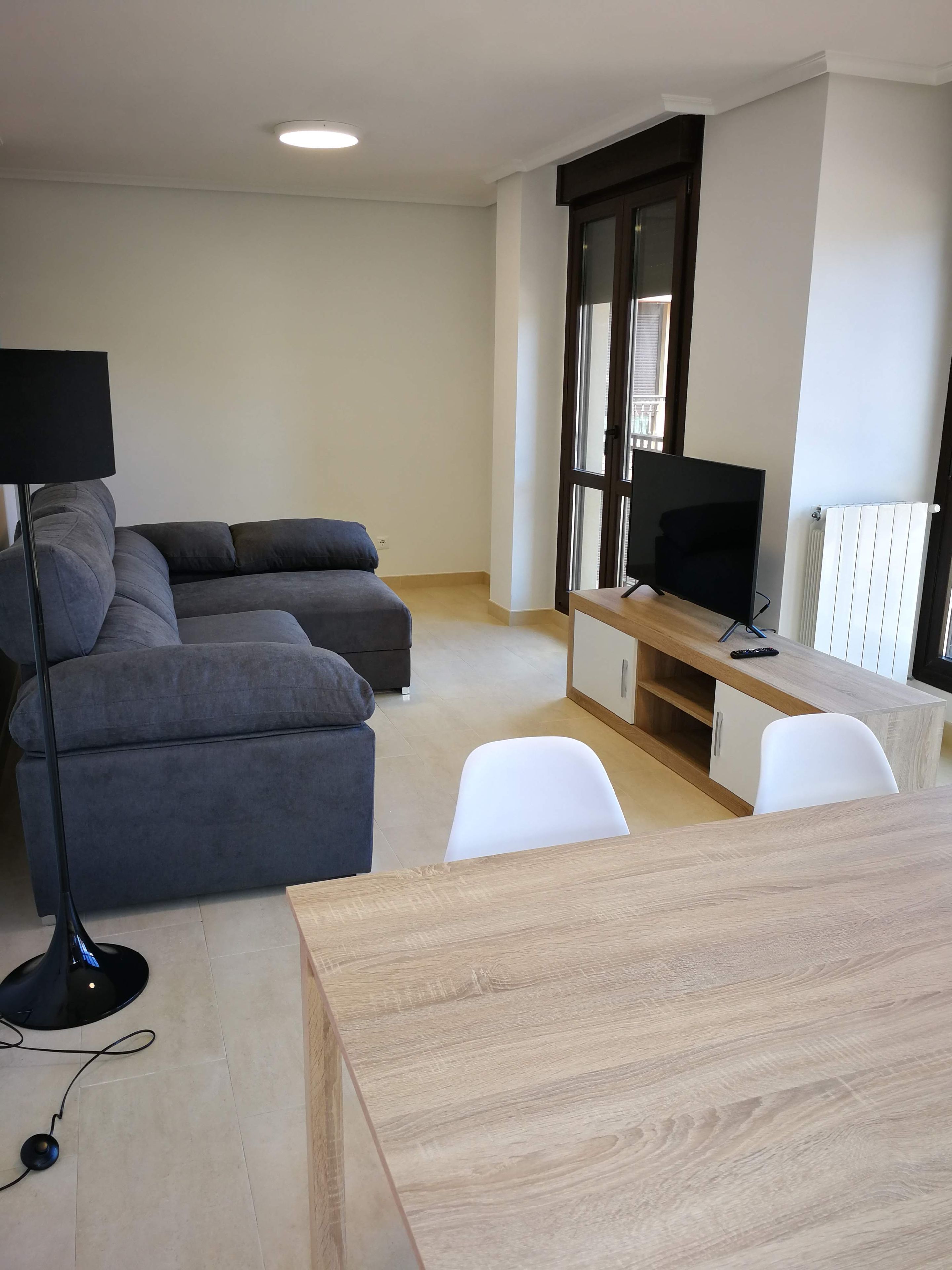 Uma sala de estar moderna com um sofá cinzento, uma mesa de centro de madeira, uma TV em um móvel e uma cadeira branca.