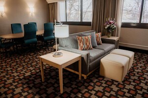 Quarto de hotel com sofá, banquinho, mesa, cadeiras e luminária.