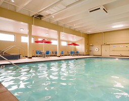 Piscina interna com água cristalina, cadeiras azuis e guarda-sóis vermelhos.