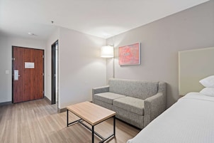 Quarto de hotel com porta de madeira, sofá cinza, mesa pequena, luminária e cama.