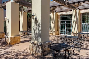 Um pátio externo com várias mesas e cadeiras, pilares de pedra e uma estrutura de pérgola.