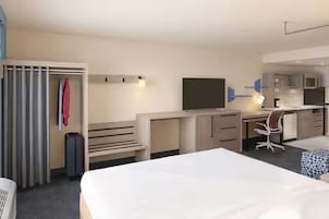 Quarto de hotel com cama, escrivaninha, cadeira e cozinha compacta.