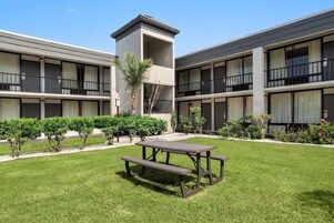 Um pátio com mesa para piquenique, cercado por sebes bem aparadas e um edifício com sacadas.