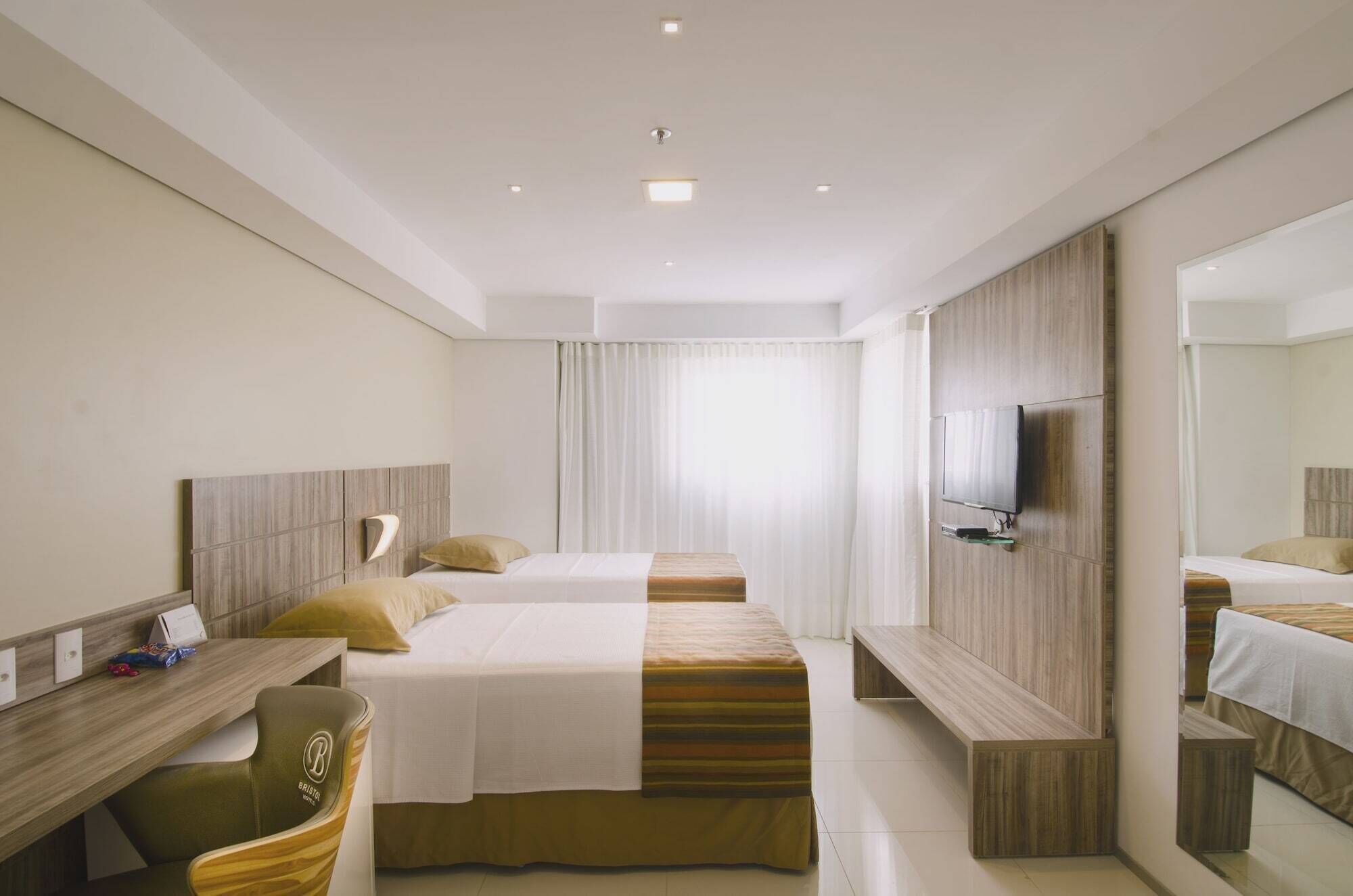 Quarto de hotel com duas camas, uma escrivaninha, uma cadeira, uma televisão e um espelho grande.