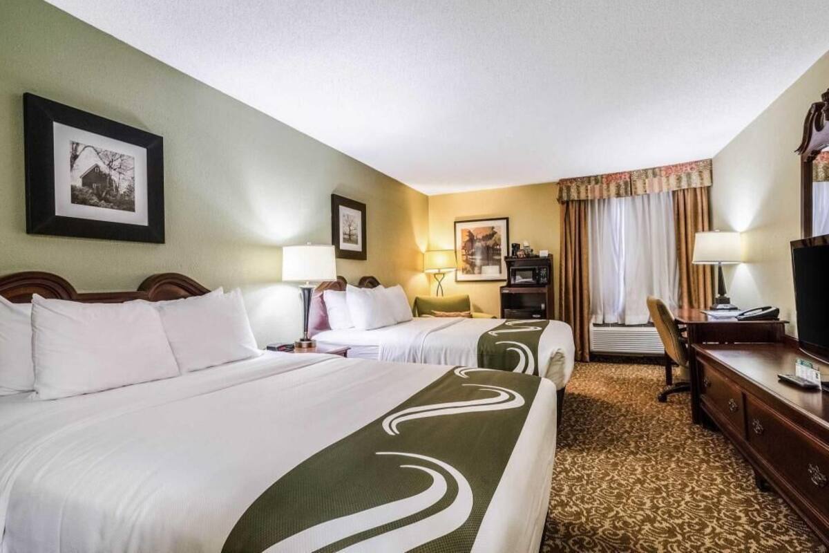 Quarto de hotel com duas camas, uma escrivaninha, uma cadeira, televisão e quadros nas paredes.