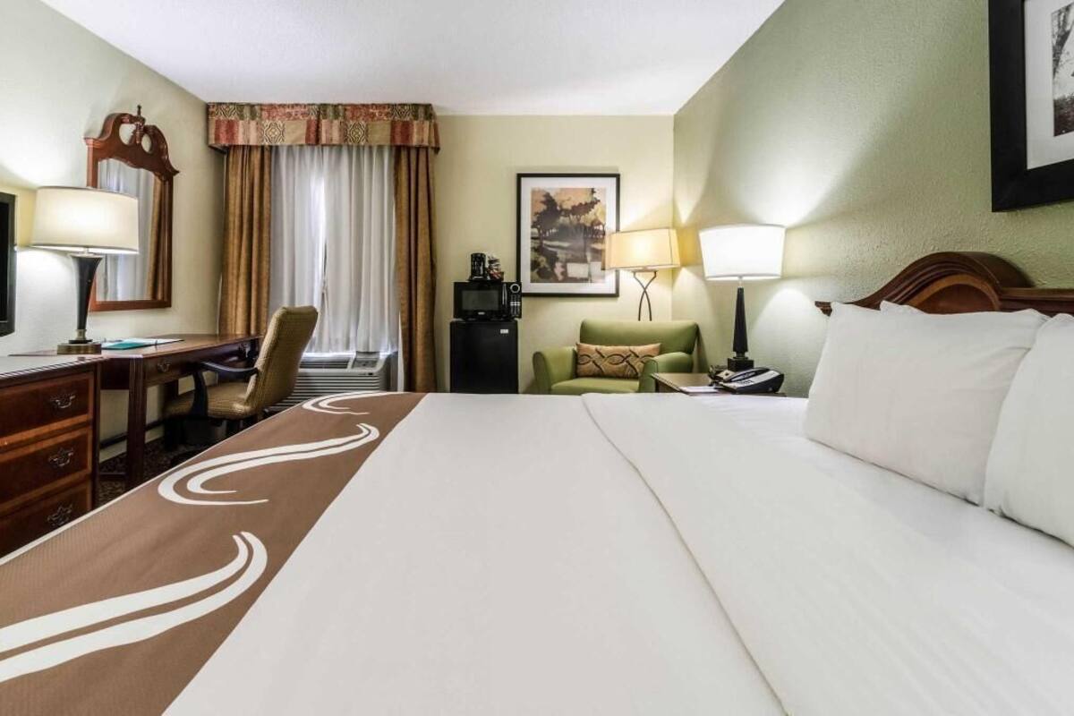 Quarto de hotel com uma cama grande, uma escrivaninha com cadeira, televisão, cafeteira e um quadro na parede.