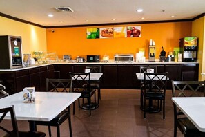 Restaurante com mesas brancas e cadeiras pretas, um balcão com exposição de comida e uma máquina de café.