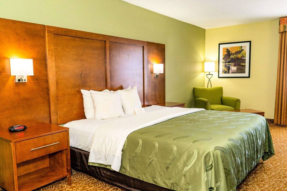 Quarto de hotel com uma cama grande, mesas de cabeceira, uma poltrona verde e um quadro na parede.