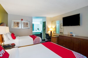 Quarto de hotel com duas camas, uma escrivaninha, uma cadeira, televisão e banheiro visível através de uma porta aberta.