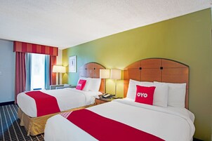 Quarto de hotel com duas camas, cada uma com colcha vermelha e branca, uma mesinha de cabeceira com abajur e uma janela com cortinas.