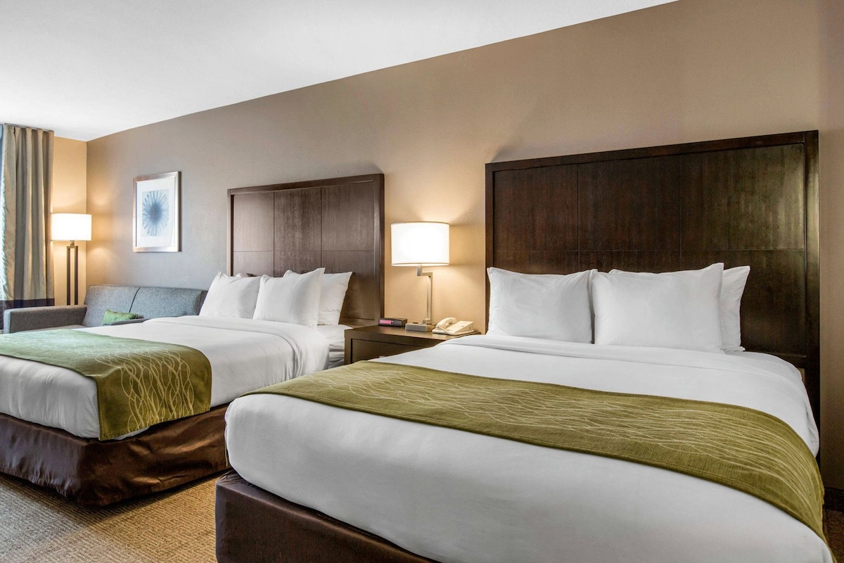 Quarto de hotel com duas camas, uma escrivaninha, uma cadeira e um abajur.