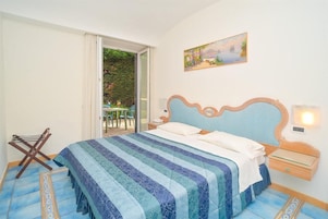 Quarto com cama, mesa de cabeceira, quadro e vista de um jardim através de uma porta aberta.