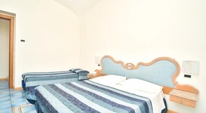 Quarto de hotel com uma cama grande, uma cama menor, uma mesa de cabeceira e uma luminária fixada na parede.