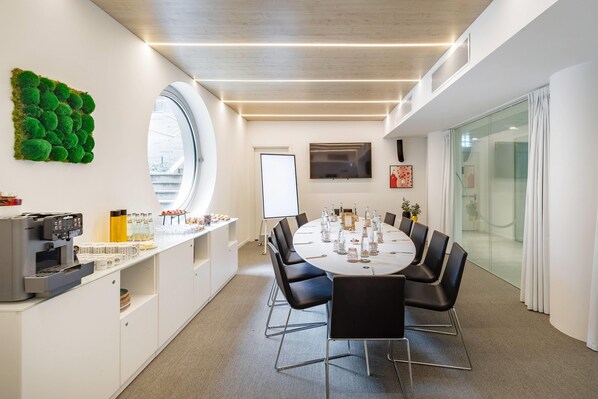 Sala de reuniões moderna com mesa redonda, cadeiras pretas, espaço para café, janela grande e decoração verde na parede.