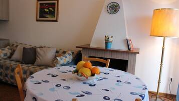 Uma sala de estar com uma mesa redonda coberta por um tecido de mesa com padrão de folha, uma tigela de frutas sobre a mesa, um sofá com capa xadrez, uma lareira com um prato decorativo e um abajur.